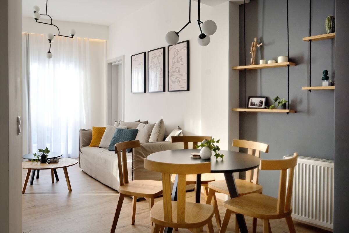 Uma sala de estar moderna com um sofá, mesas redondas de madeira e cadeiras. Há quadros nas paredes e prateleiras com objetos decorativos.