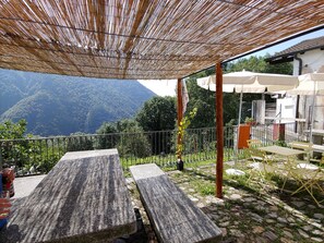 Uma área externa com mesas e cadeiras de pedra, um toldo de bambu e vista para uma paisagem montanhosa.