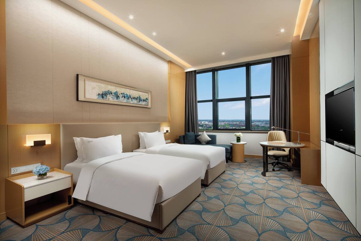 Quarto de hotel com duas camas, uma escrivaninha, uma cadeira, televisão e uma grande janela com vista para a cidade.