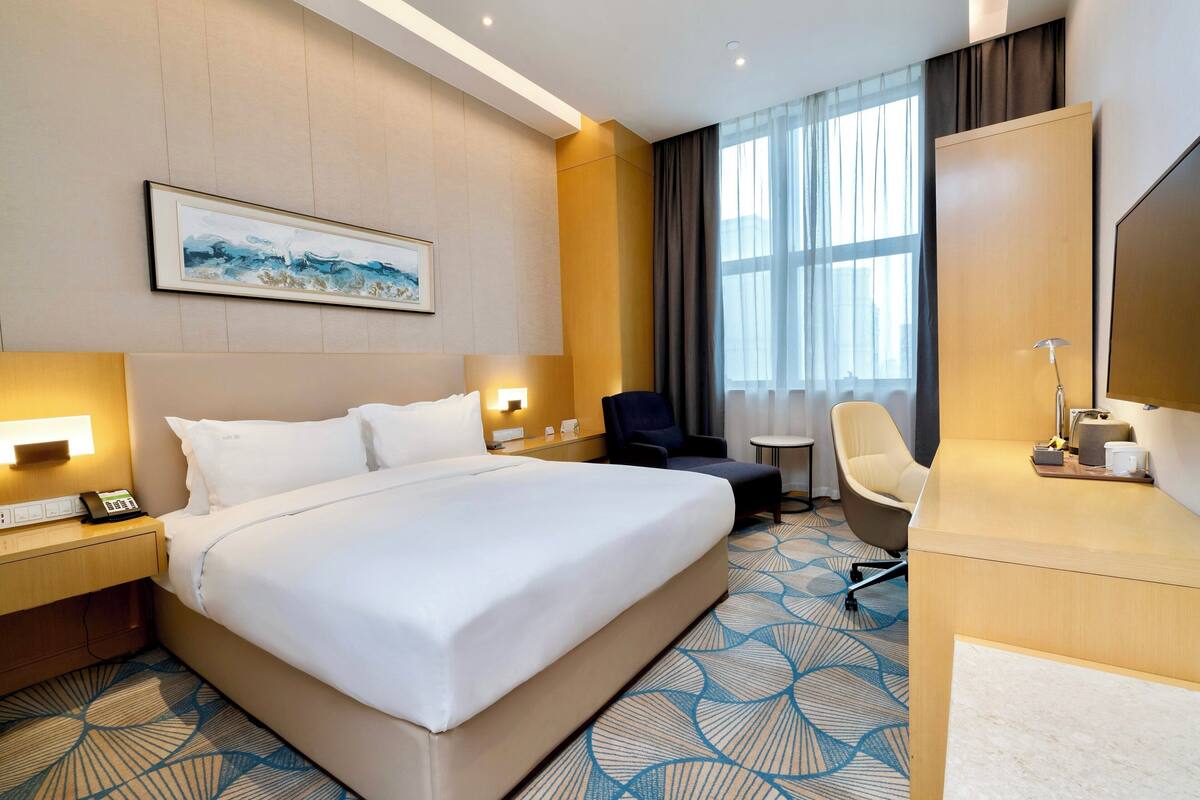 Quarto de hotel com uma cama grande, mesas de cabeceira, escrivaninha, cadeira e uma janela ampla com cortinas.