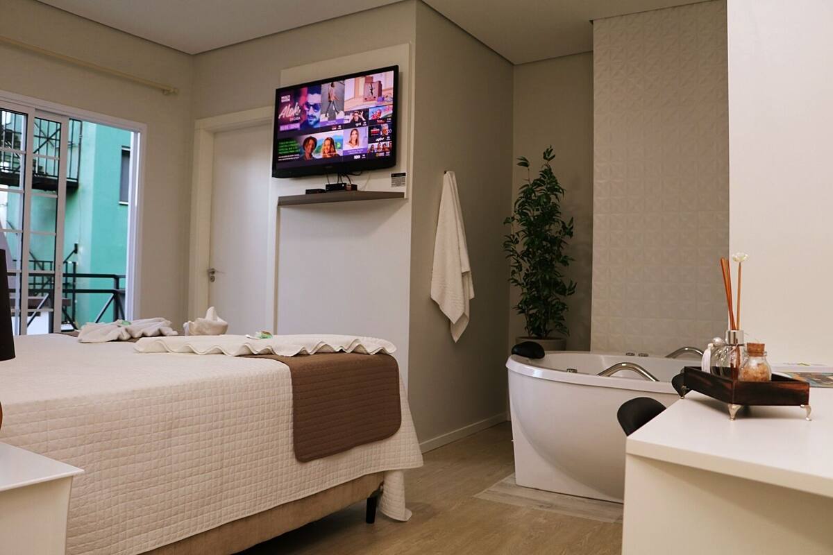 Quarto de hotel com cama, televisão fixada na parede, banheira e planta.