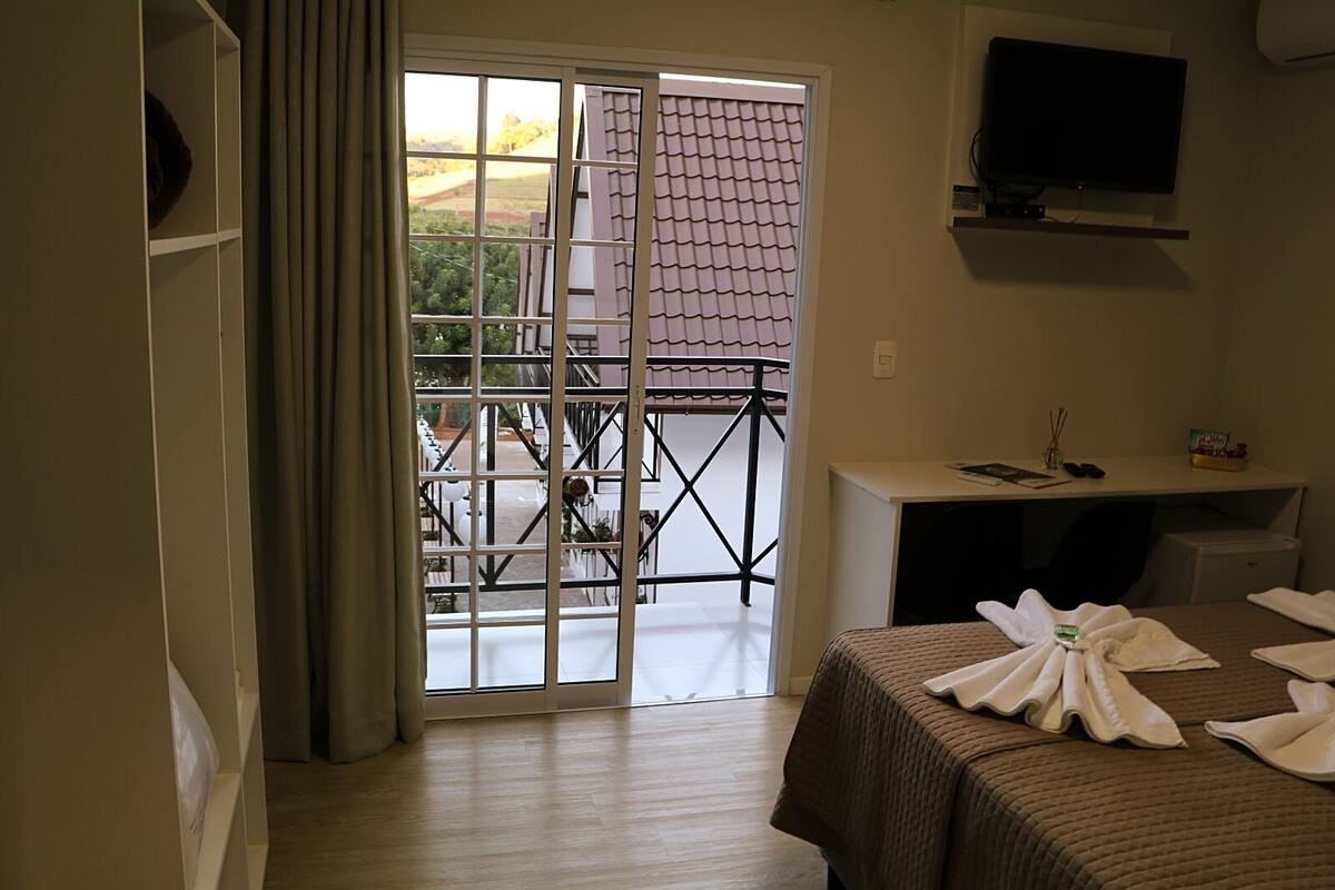Quarto de hotel com cama, escrivaninha, televisão e varanda com vista para o campo.