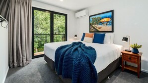 Quarto moderno com uma cama grande, uma mesa de cabeceira com abajur, um quadro de uma praia e uma janela com cortinas.