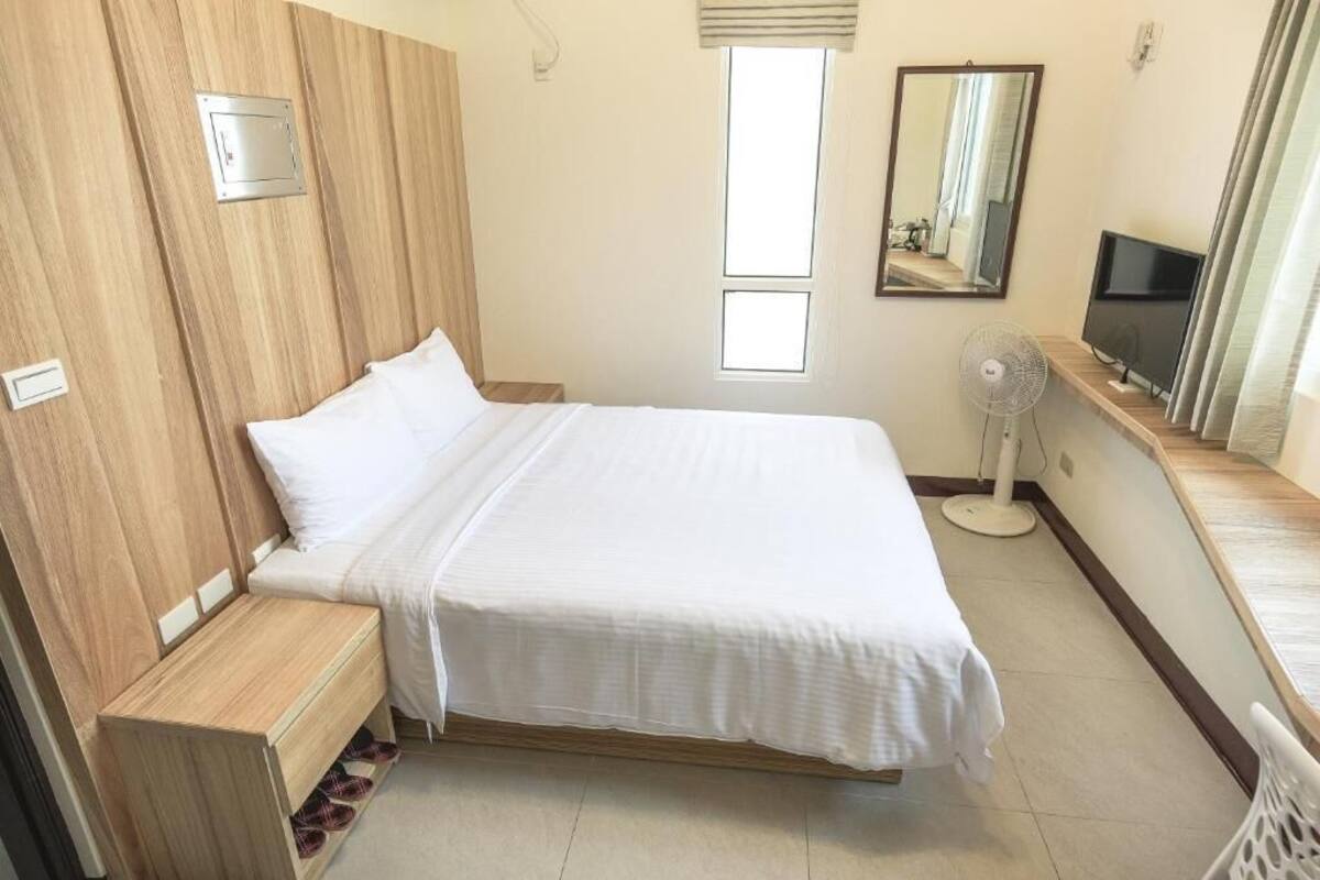 Quarto bem organizado com cama, mesa de cabeceira, um pequeno ventilador, TV e janela.