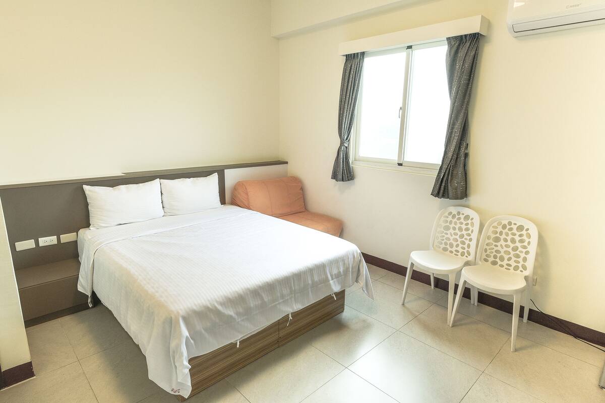 Quarto com cama, duas cadeiras, janela com cortinas e ar condicionado.