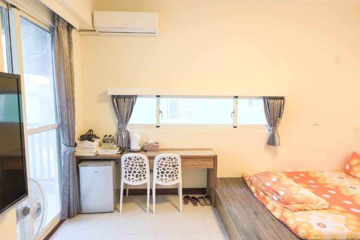 Quarto compacto com cama, escrivaninha com duas cadeiras, mini-geladeira e televisão.