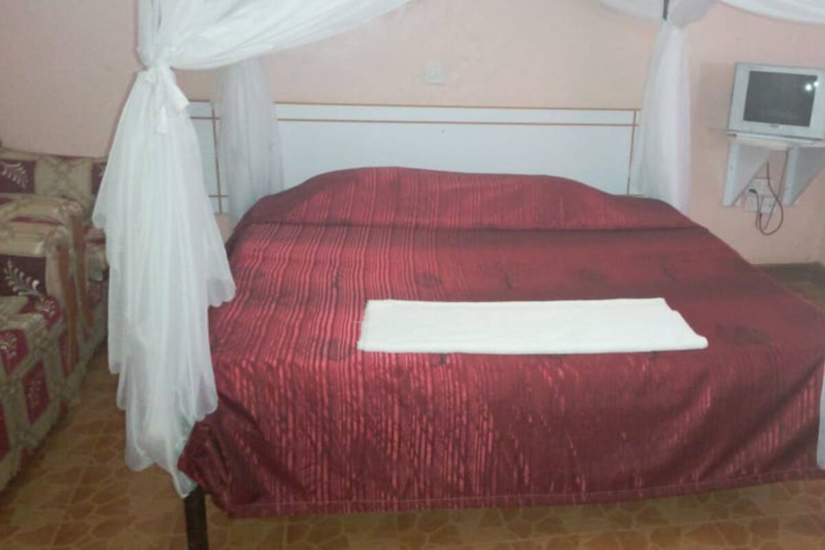 Quarto com cama coberta por um edredom vermelho e cortinas brancas. Há uma televisão sobre uma mesinha ao lado da cama.