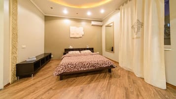 Quarto com piso de madeira, cama com colcha estampada, mesa de cabeceira, espelho e ar-condicionado fixado na parede.