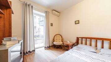 Quarto de hotel com uma cama, uma cadeira, uma mesinha e uma janela com cortinas.