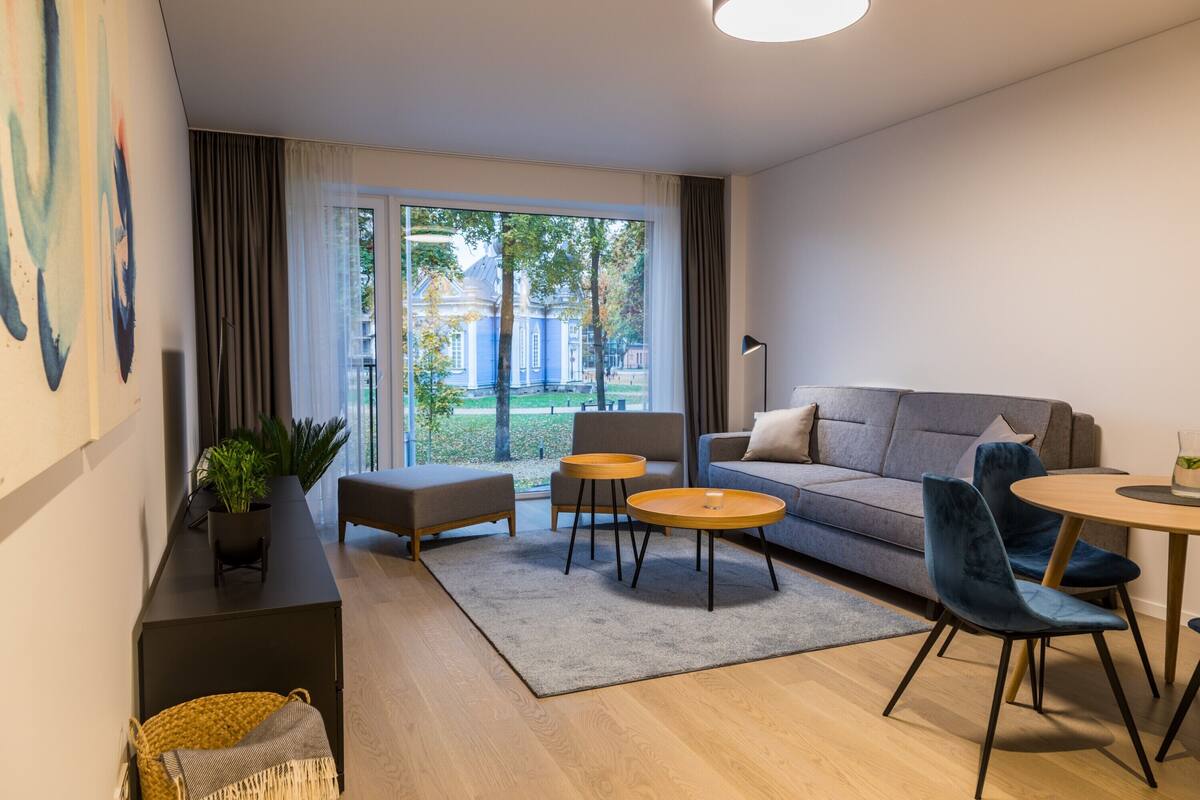 Uma sala de estar moderna com sofá, duas mesas redondas, um banco e uma janela grande com vista para edifícios e árvores.