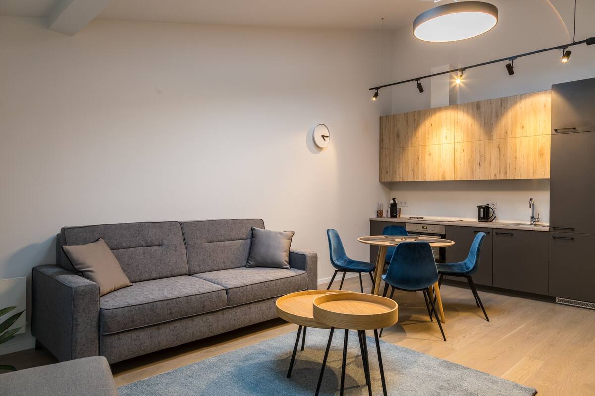 Uma sala de estar moderna com um sofá cinzento, cadeiras azuis, uma pequena mesa de madeira e uma área de cozinha com armários de madeira e um candeeiro redondo.