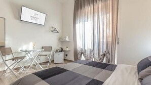 Quarto de hotel com cama, escrivaninha, cadeira, mesa pequena e televisão fixada na parede.