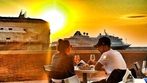 Um casal jantando à mesa com vista para um navio de cruzeiro e um edifício grande ao fundo.