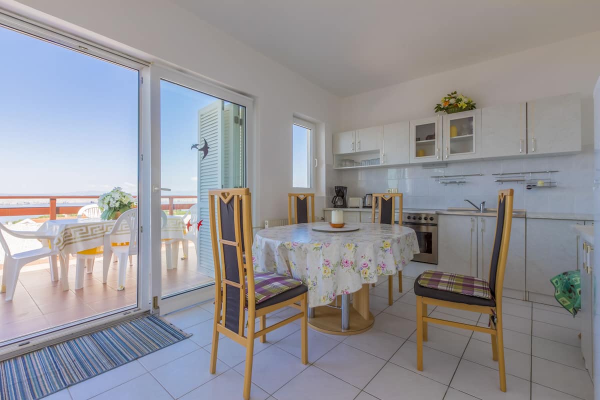 Uma área de refeições com mesa e cadeiras, uma cozinha com armários e eletrodomésticos, e uma porta de vidro deslizante que dá para uma varanda.