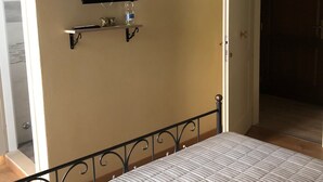 Quarto com cama, televisão montada na parede, prateleira com uma garrafa de água e detalhe decorativo na janela.