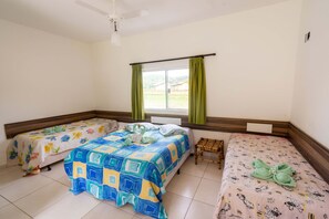 Quarto com duas camas, janela com cortinas verdes, ventilador de teto e vista para uma casa.