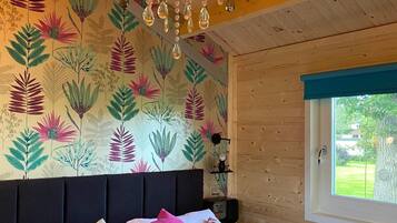 Quarto com teto de madeira, cama grande com várias almofadas, um lustre e janela com vista para vegetação.