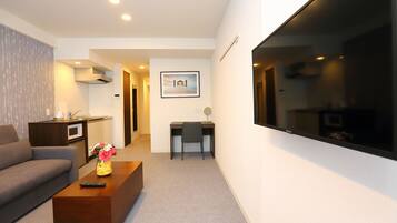 Uma sala de estar moderna com sofá, mesa de centro e televisão. Há uma escrivaninha com cadeira e um quadro na parede.