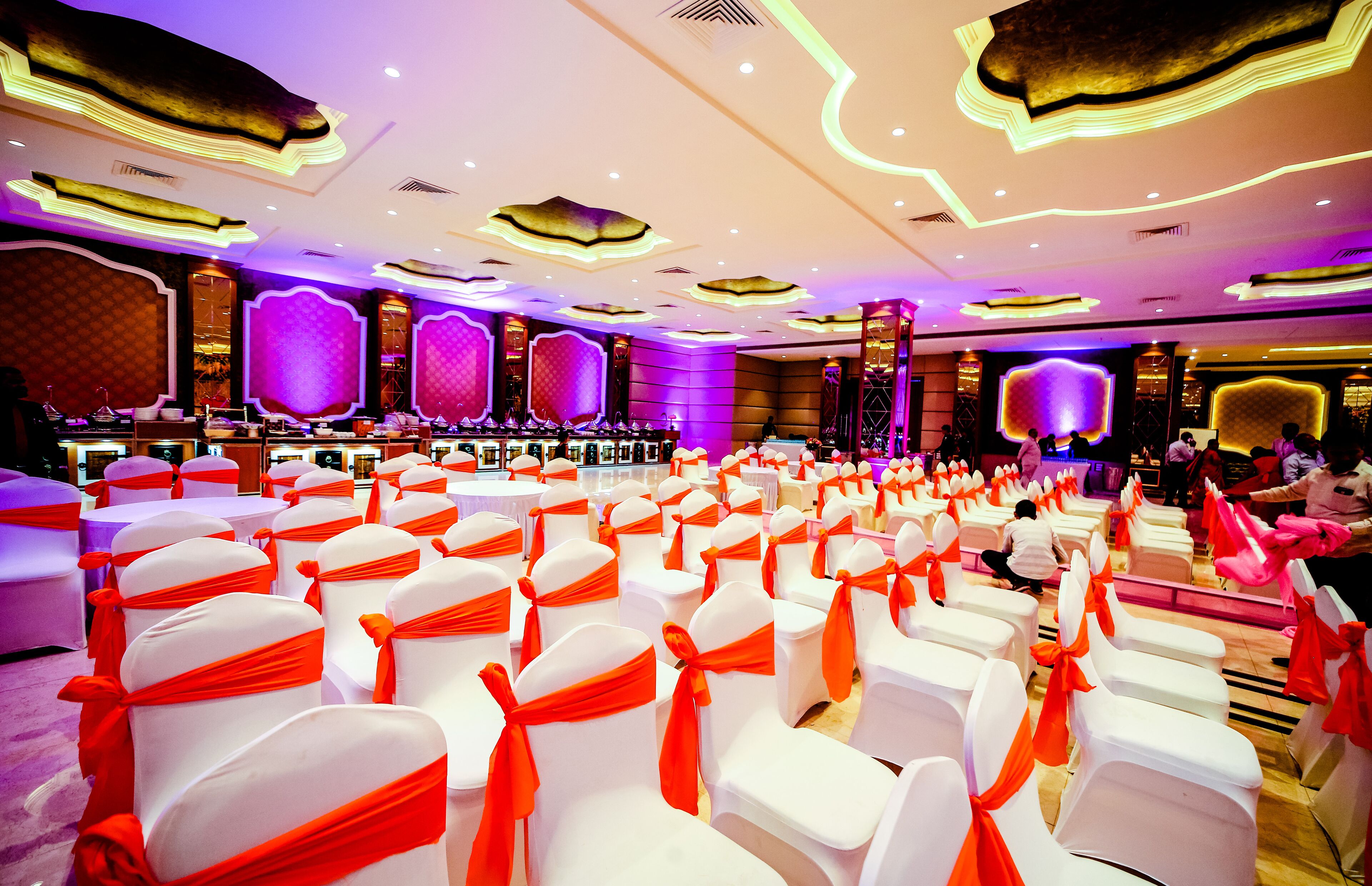 Salão de banquetes com fileiras de cadeiras brancas e fitas laranja, palco com uma tela grande e área de bar.