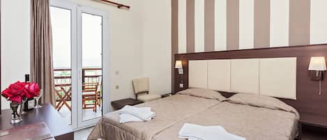 Quarto de hotel com uma cama grande, uma escrivaninha com um vaso de flores, uma cadeira e vista para o exterior por uma porta de correr.