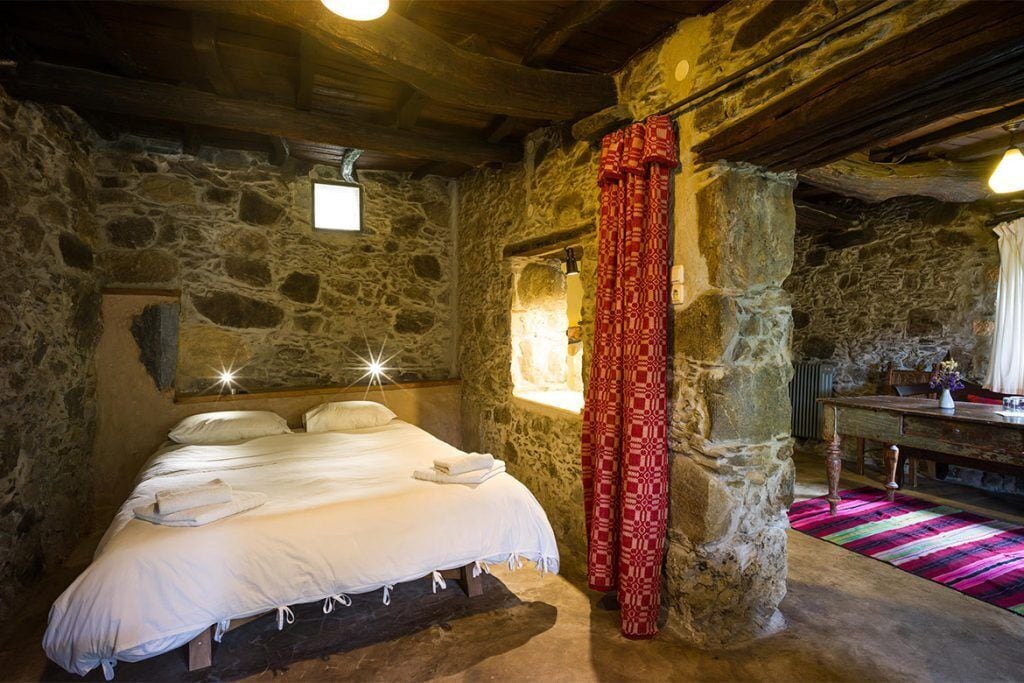 Um quarto com paredes de pedra, uma cama, um banco de madeira, uma mesa com um vaso e um tapete colorido.