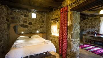 Um quarto com paredes de pedra, uma cama, um banco de madeira, uma mesa com um vaso e um tapete colorido.