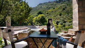 Uma mesa com uma garrafa, uma tigela de frutas e uma taça de vinho, tudo sobre um terraço com vista para uma paisagem montanhosa.