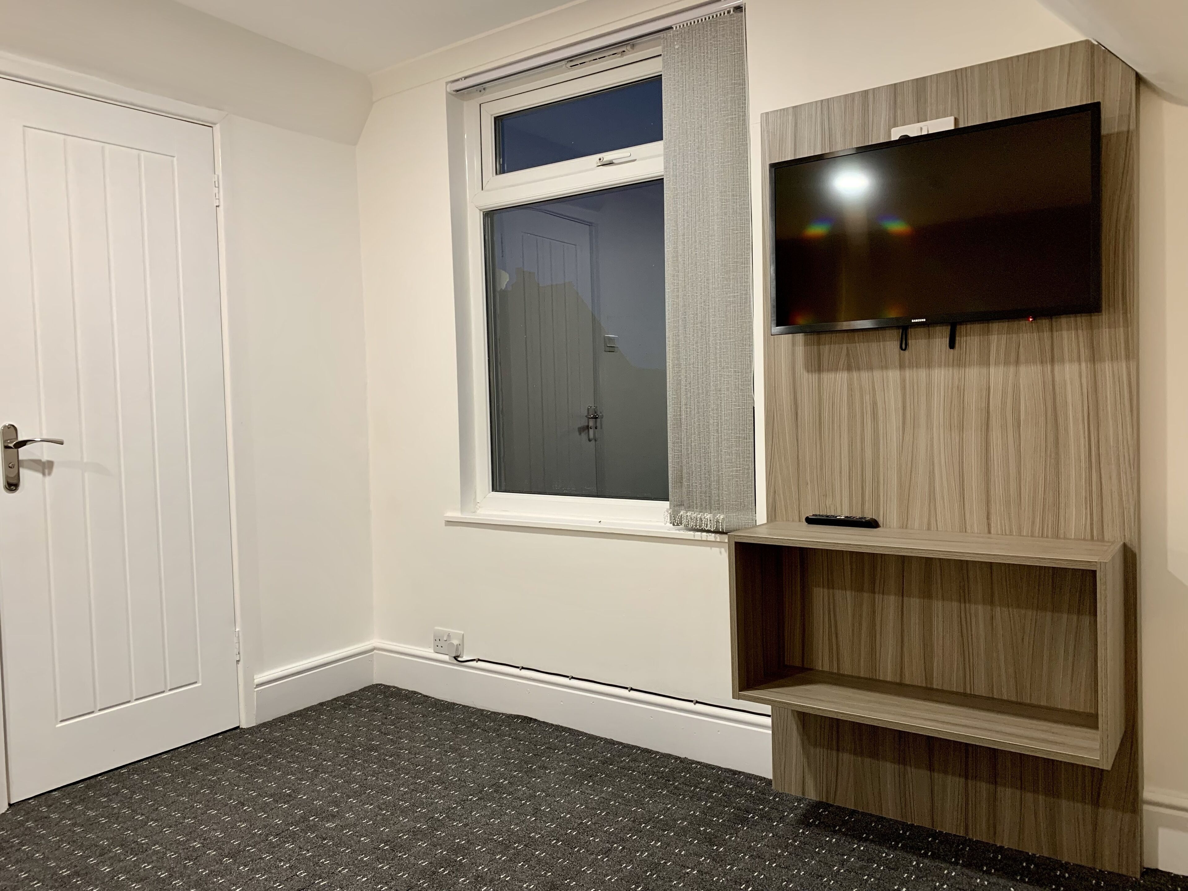 Quarto com porta fechada, janela com cortina, televisão de tela plana fixada em parede de madeira e piso coberto por carpete.