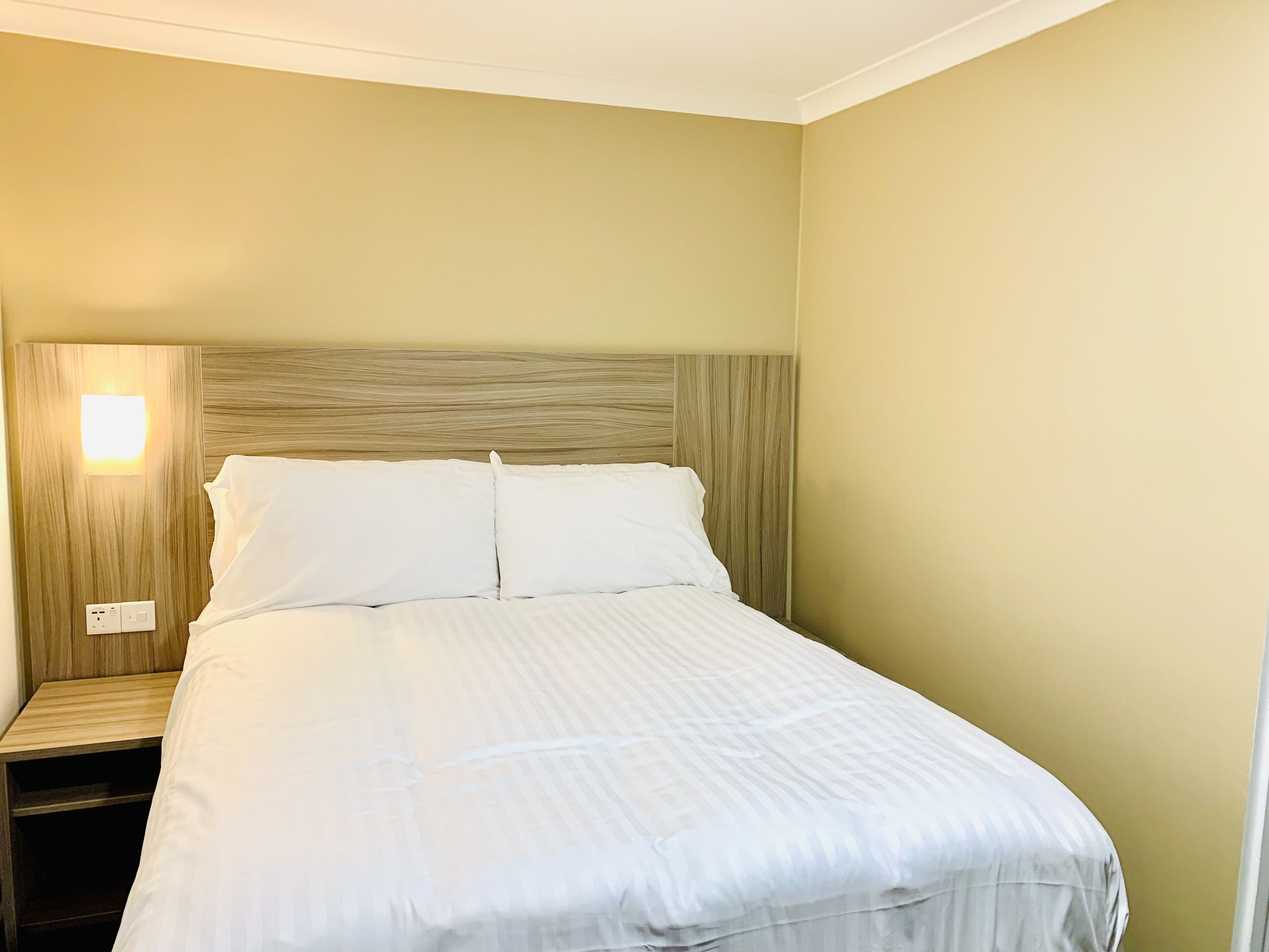 Cama bem arrumada com lençóis brancos num quarto com paredes bege e cabeceira de madeira.