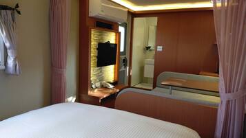 Quarto de hotel com cama, televisão presa a uma parede de pedra, espelho e banheiro visível por uma porta aberta.