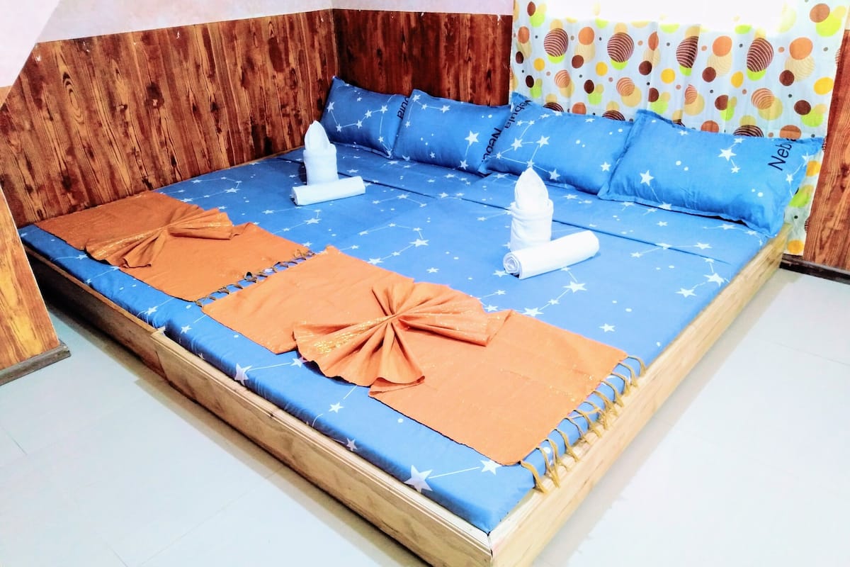 Uma cama com colcha estampada em estrelas azuis, almofadas decorativas cor de laranja e cabeceira de madeira.
