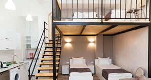 Quarto de hotel pequeno com beliche, cozinha compacta e escada que leva a um andar superior.