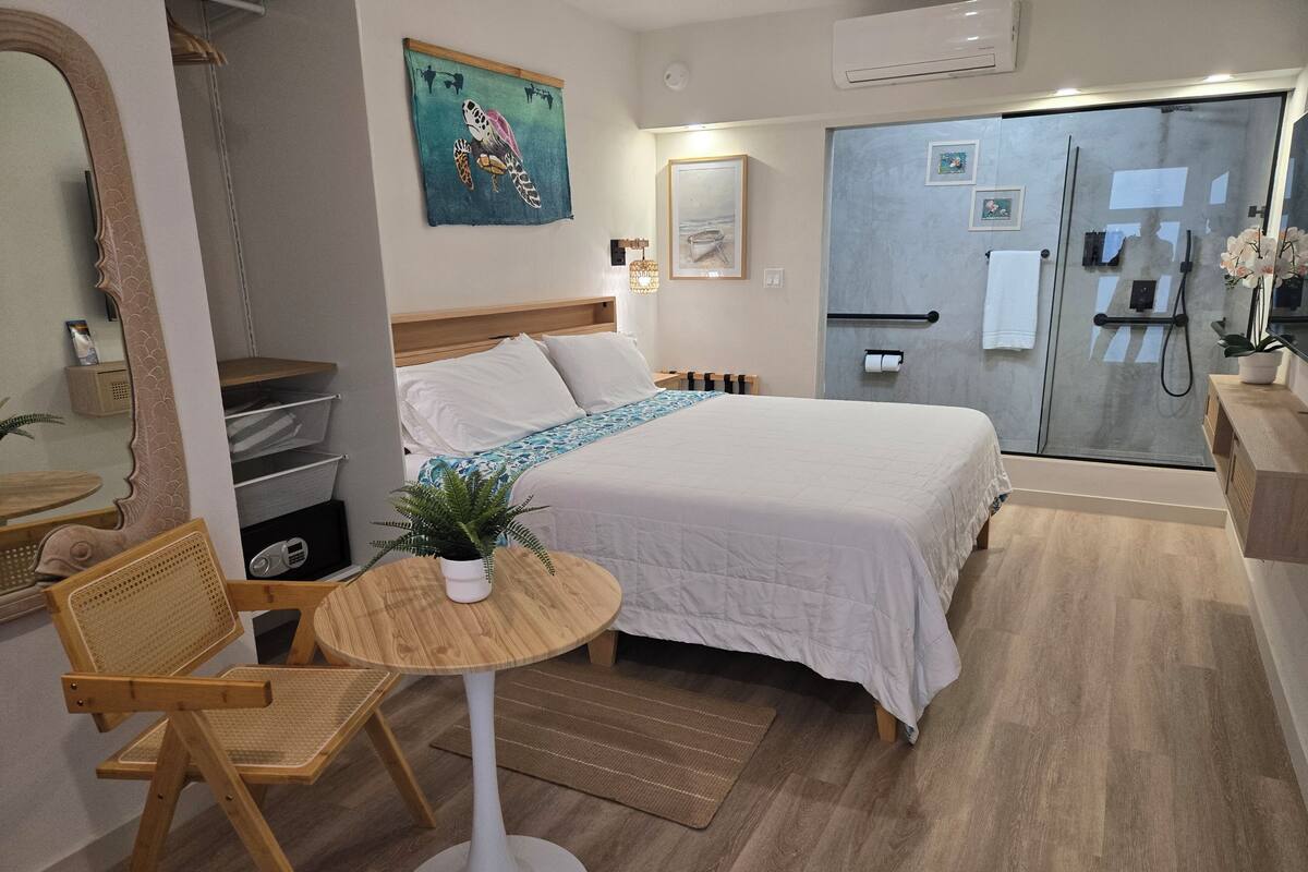 Quarto de hotel moderno com uma cama, uma mesa redonda com uma planta, uma cadeira, um espelho grande e um banheiro com porta de vidro.