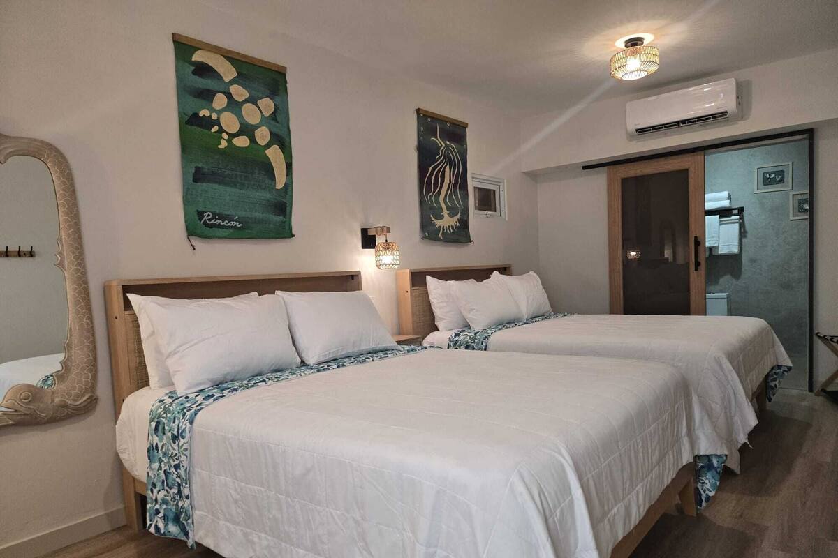Quarto de hotel com duas camas, um espelho, obras de arte, aparelho de ar condicionado e porta de acesso a um banheiro.