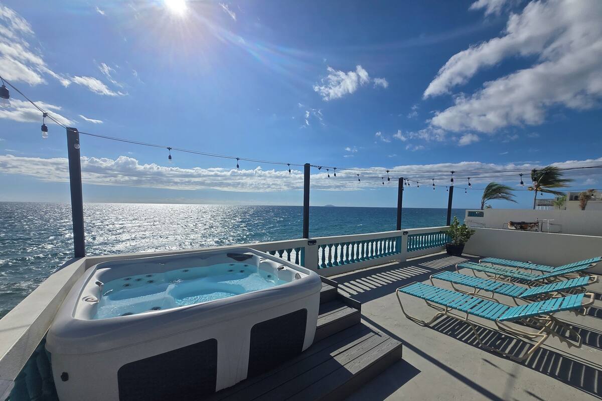 Uma banheira de hidromassagem em um deck com vista para o oceano, cadeiras de descanso e luzes de festa.