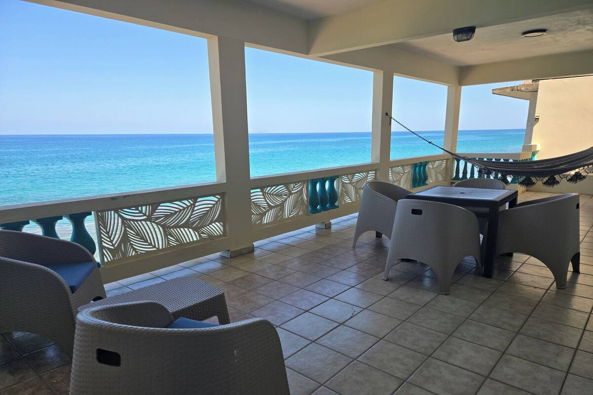 Área externa coberta com vista para o mar, com uma mesa, cadeiras, uma rede e guarda-corpo estampado.