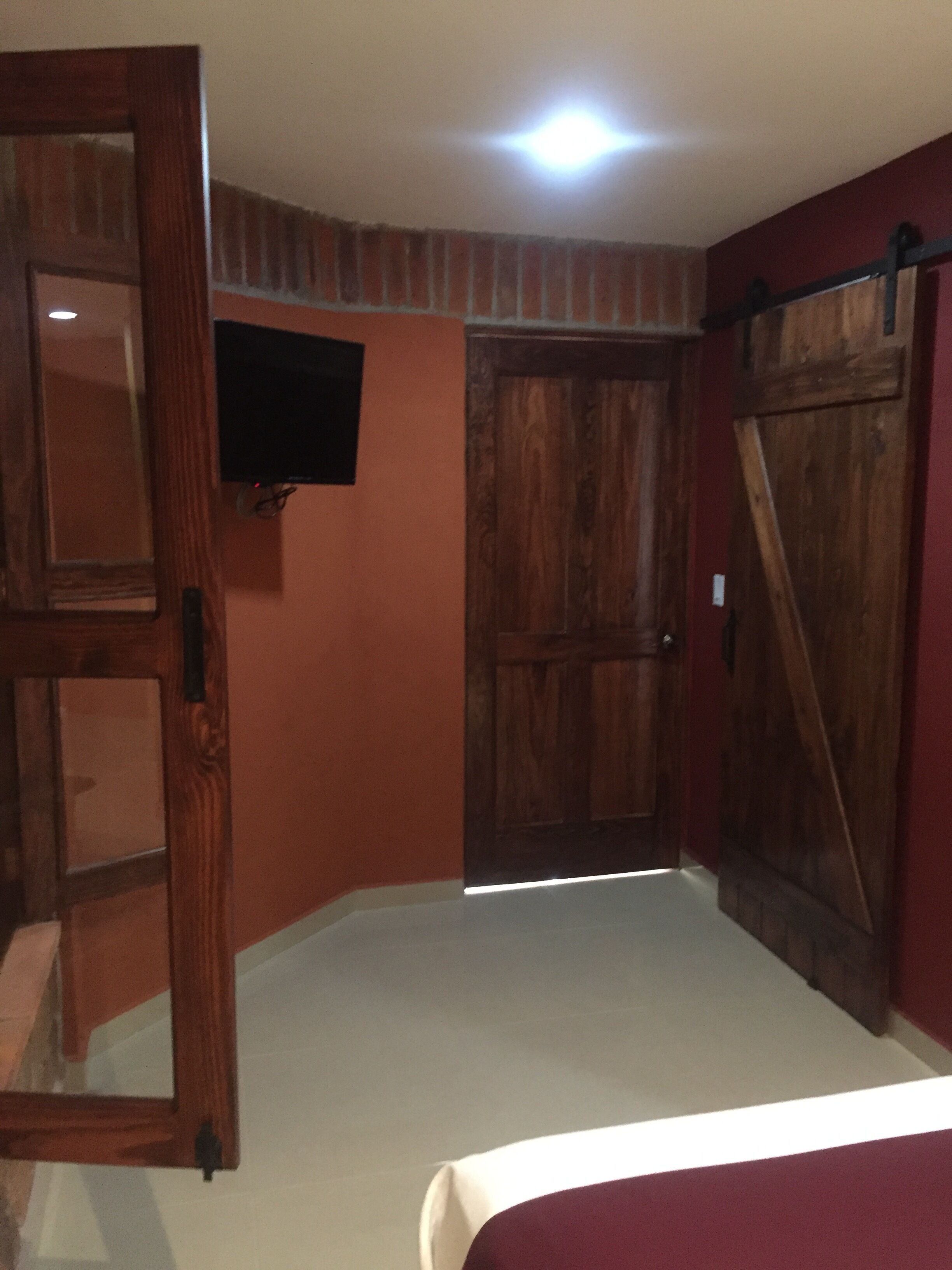 Quarto com porta de madeira, televisão fixada na parede e cama com colcha vermelha.
