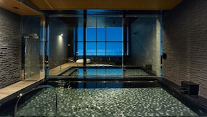 Piscina interior moderna com vista clara do oceano através de grandes janelas, teto de madeira e paredes de pedra.