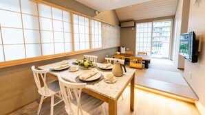 Uma sala de jantar com mesa de madeira e cadeiras brancas, uma pequena cozinha e uma porta deslizante para uma varanda.