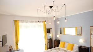 Quarto de hotel moderno com uma cama grande, banheiro separado com banheira, piso de madeira e um espelho grande sobre a cama.
