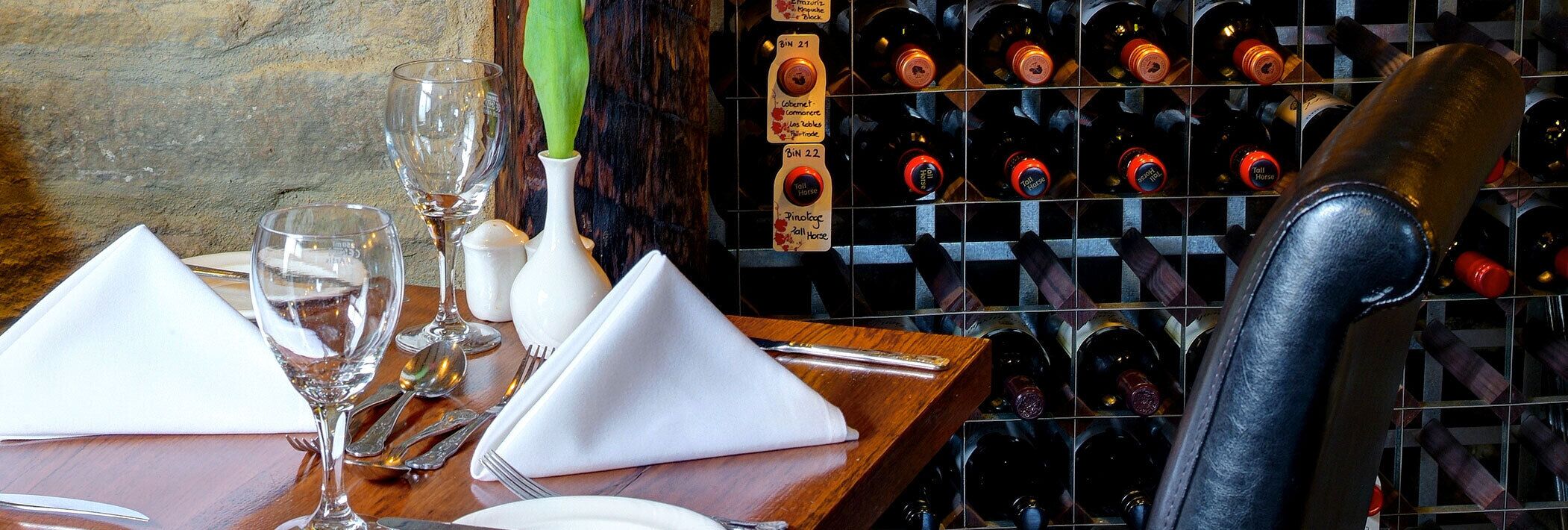 Uma mesa de jantar posta com guardanapos brancos, talheres e copos de vinho, com um suporte de vinhos ao fundo.