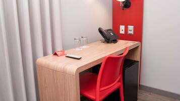 Um espaço de trabalho pequeno com uma mesa, uma cadeira vermelha, um cesto de lixo preto e um telefone fixado na parede.