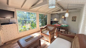Quarto com televisão, mobiliário de vime, mesa de jantar e vista para o verde lá fora.