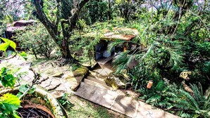 Um deck de madeira com um gazebo rústico, rodeado por folhagem tropical densa e um caminho de pedra.