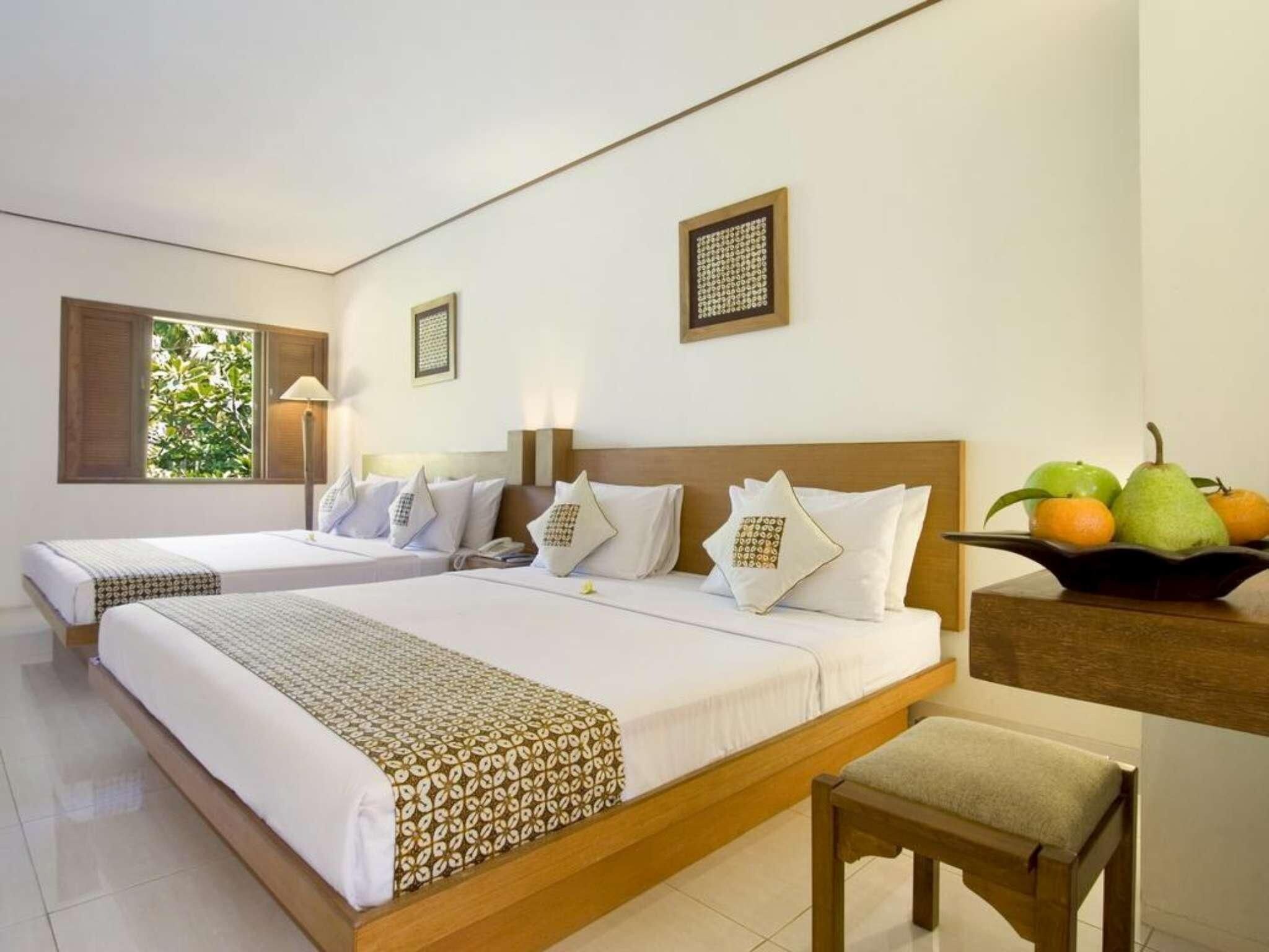 Quarto de hotel com duas camas, uma escrivaninha com frutas e janela com vista para o verde.