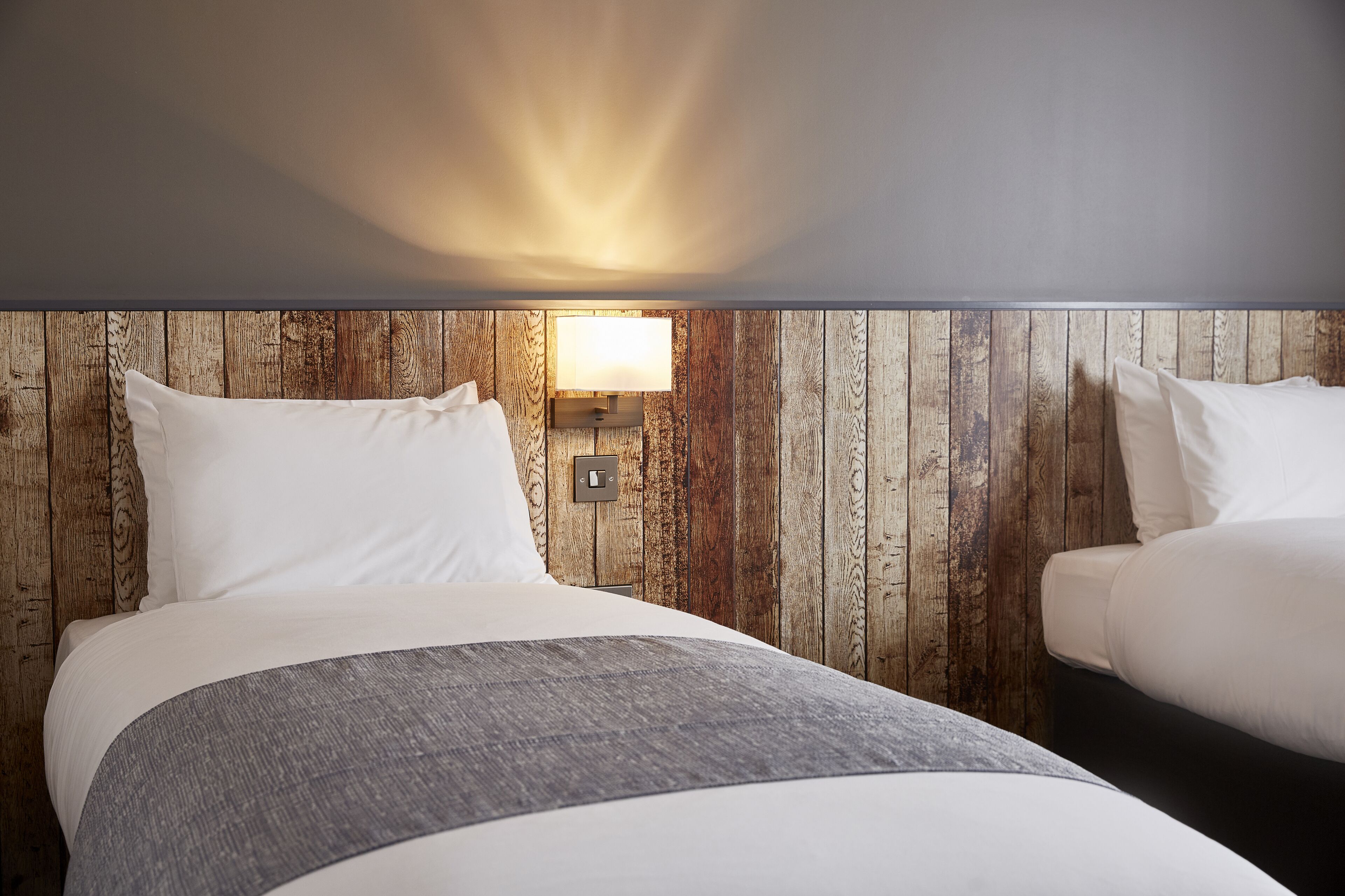 Quarto de hotel com parede de madeira, cama com roupa de cama branca e luminária de cabeceira.
