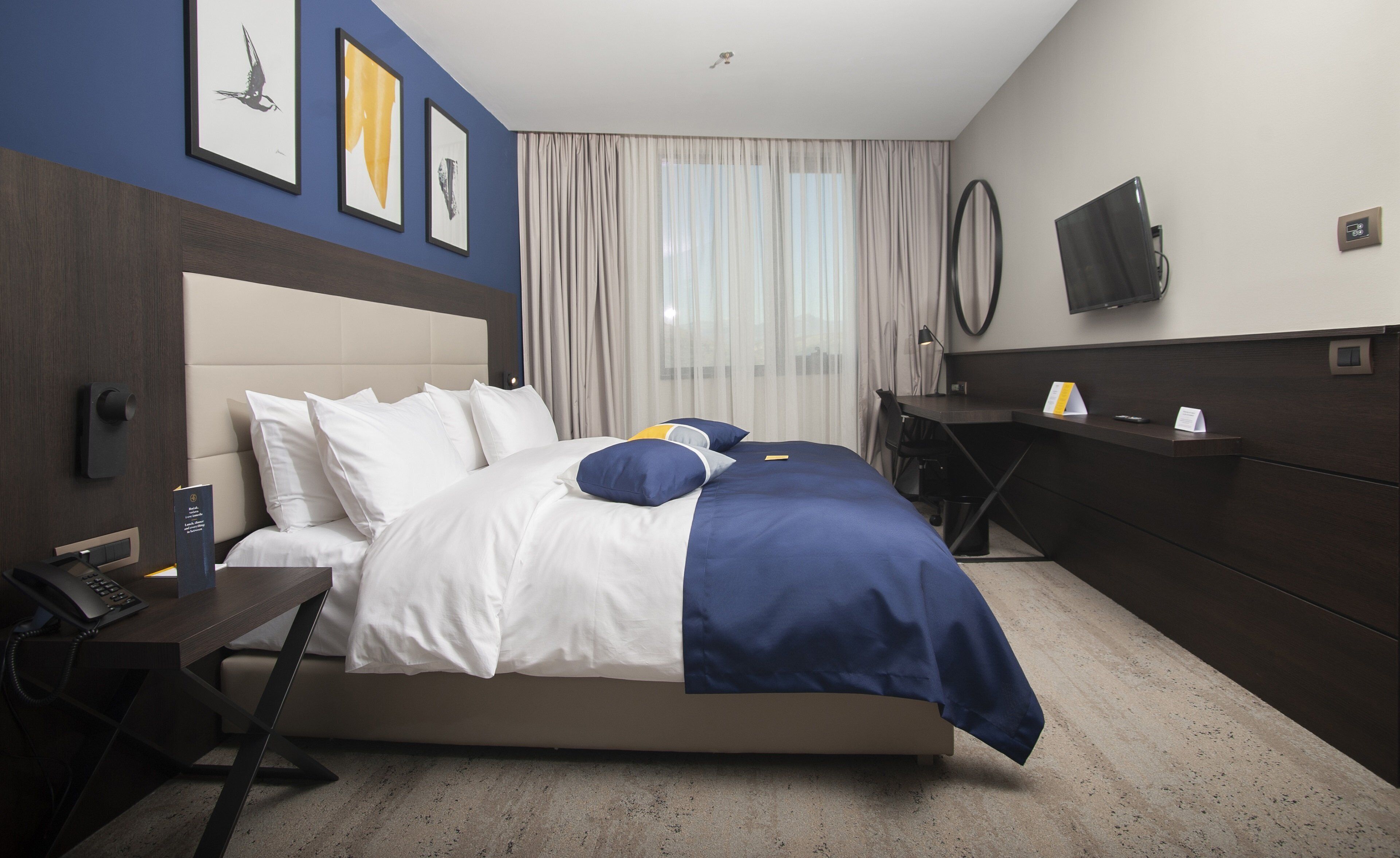 Quarto de hotel com cama, escrivaninha, telefone, televisão e janela com cortinas.