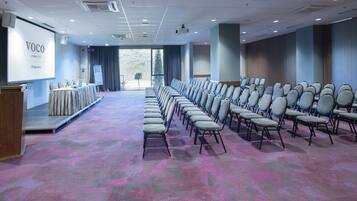Uma sala de conferências com fileiras de cadeiras, um púlpito e uma tela de projeção exibindo 'VOCO AN IHG HOTEL Podgorica'.