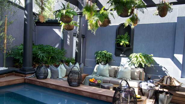 Uma área na beira da piscina com deck de madeira, um sofá com almofadas, vasos suspensos e uma mesa pequena com frutas e uma vela.