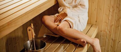 Uma pessoa sentada em um banco de madeira em um salão de sauna, com um balde de metal e uma escova de madeira.